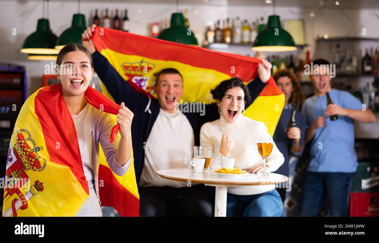 Company of excited young adult sports fans waving flag of Spain and ...