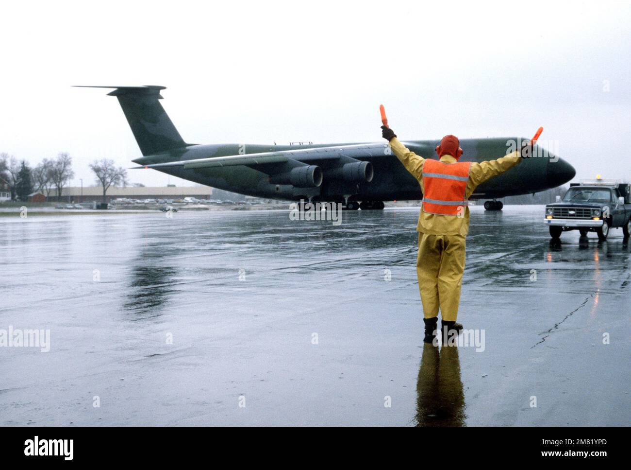 A ground crewman wearing foul weather gear uses flashlights to signal a ...