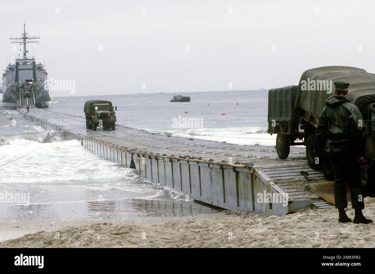 M-809 5-ton trucks are driven from a tank landing ship across a pontoon ...
