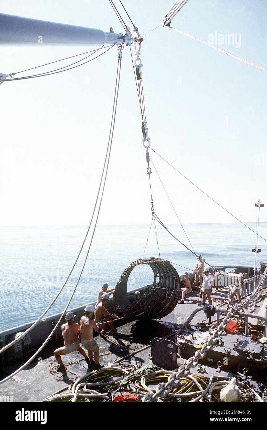 Crewmen use a crane to lift a piece of a rocket engine aboard the ...