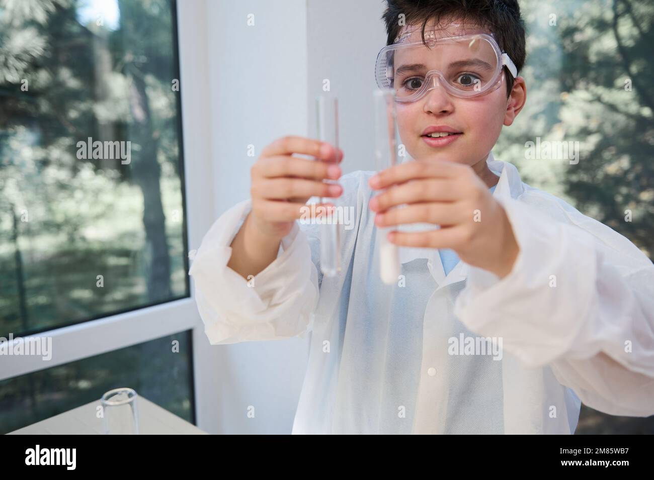 Smart schoolboy with safety goggles, in lab coat, looking at test tubes ...