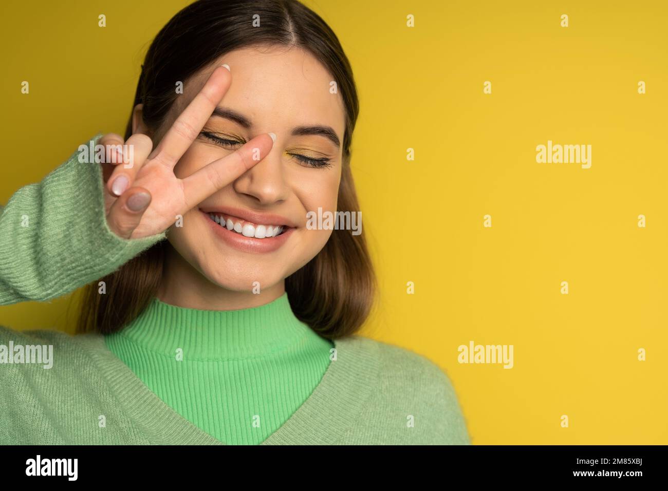 Positive teen girl in jumper showing peace sign on yellow background ...