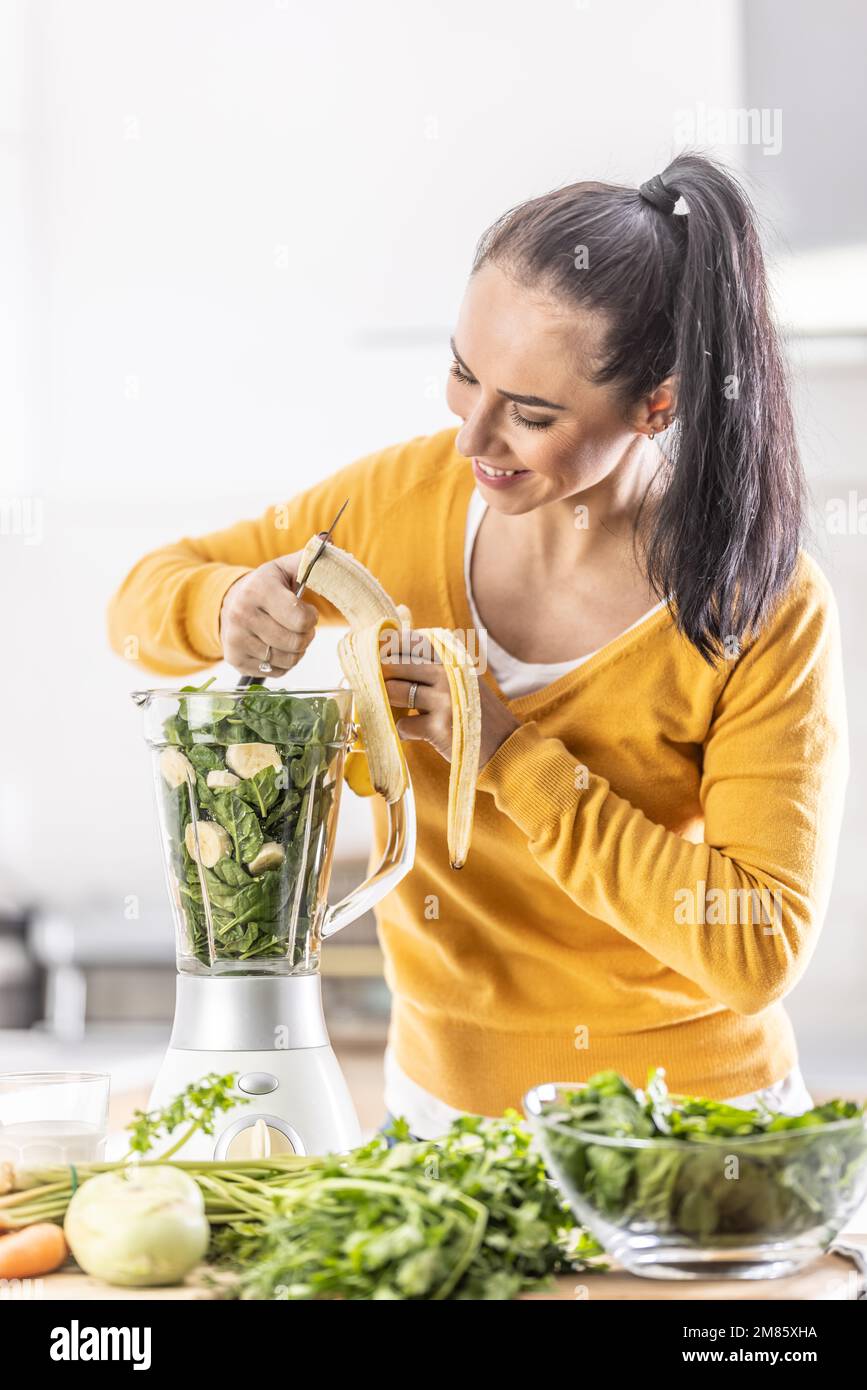 A Woman In The Kitchen Cuts A Banana In A Blender Together With Spinach