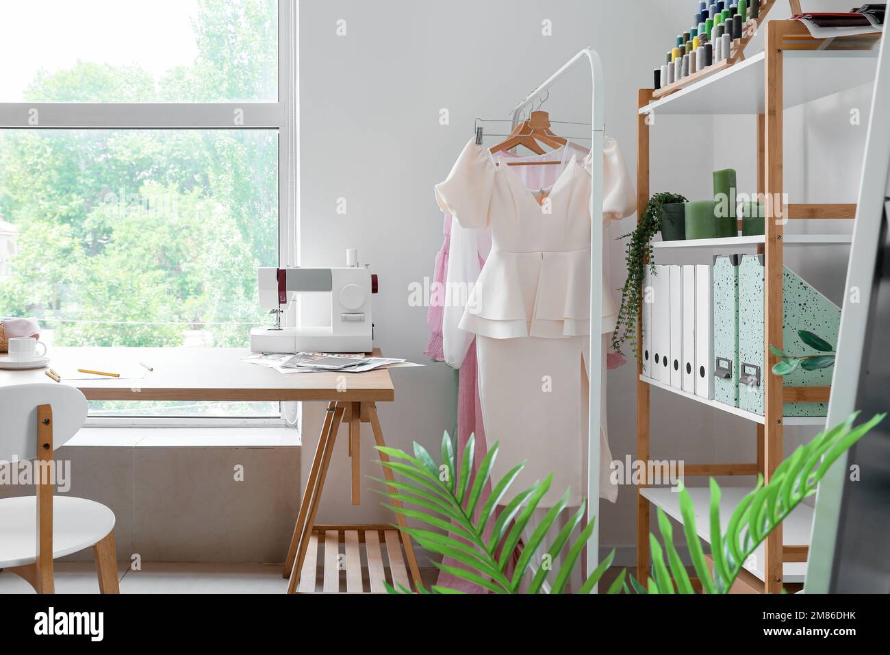 Interior of light atelier with tailor's workplace, clothes and shelving ...