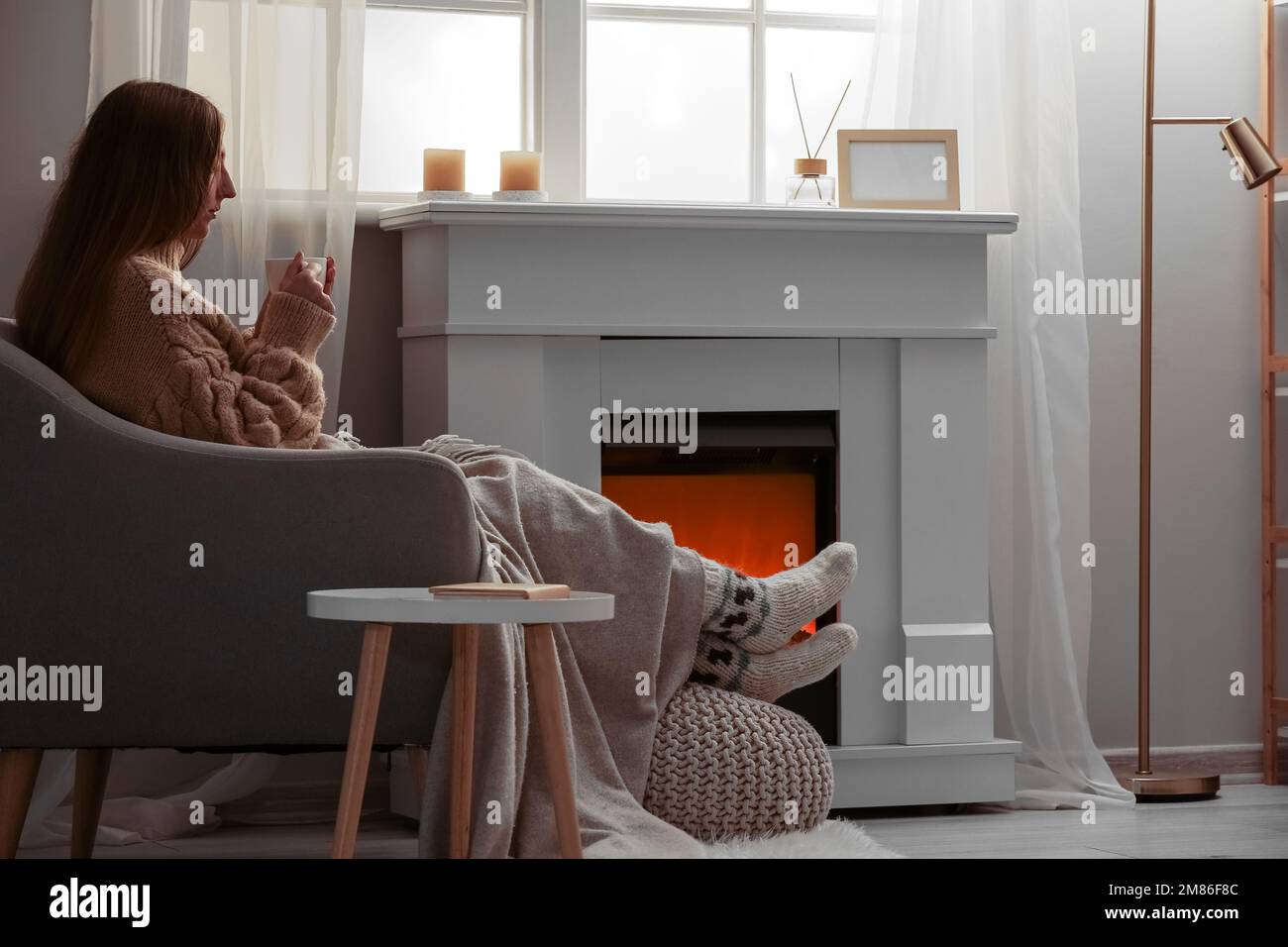Young woman with cup of tea sitting in armchair near fireplace at home ...