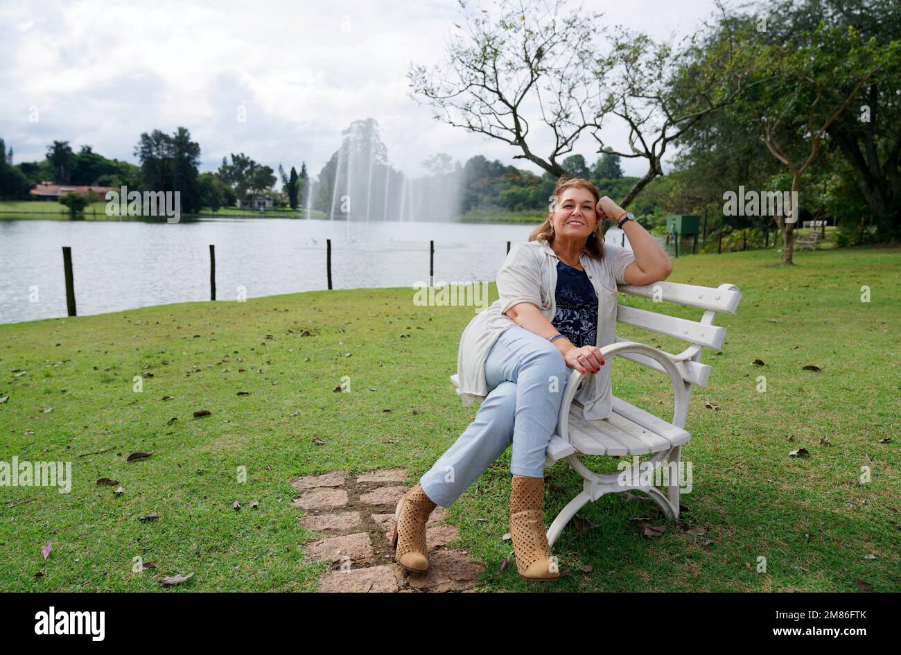 A caucasian woman sitting on a white wooden bench in a park with a lake ...