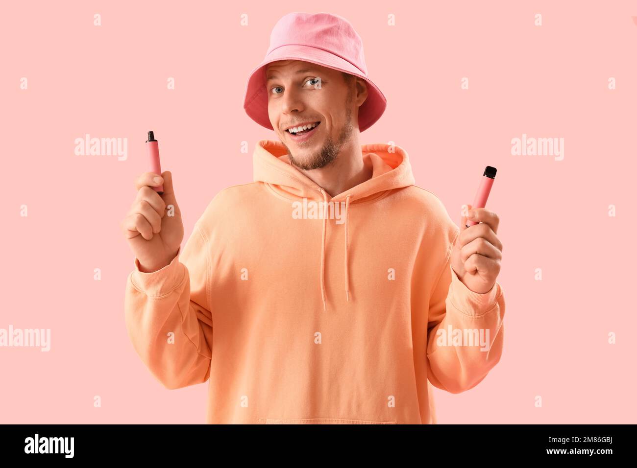 Young man in bucket hat with disposable electronic cigarettes on pink ...