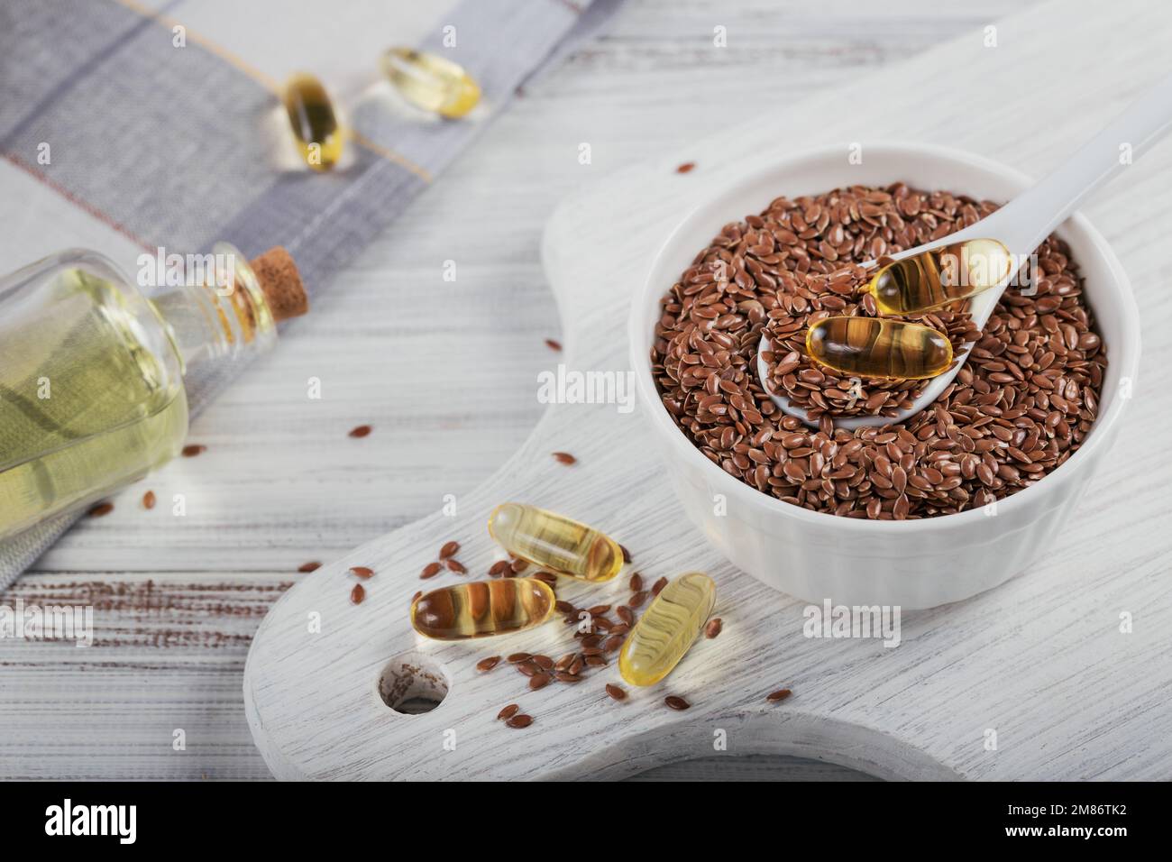 Brown flax seed or linseed in small bowl and gelatin capsules with omega oil on a white wooden ...