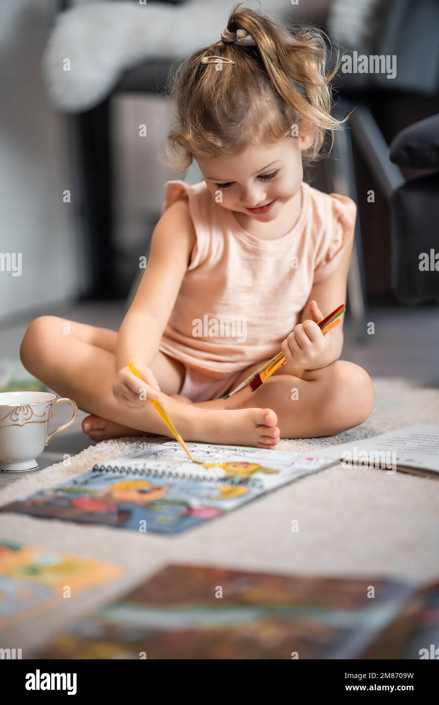 Little girl sits on the floor at home and draws with paints and brushes ...