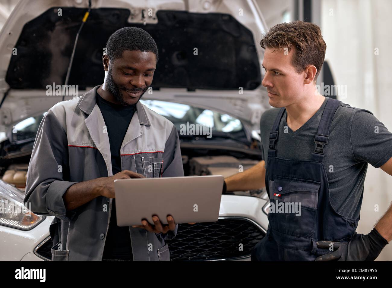 Handsome diverse mechanics in uniform examining car, using laptop while ...