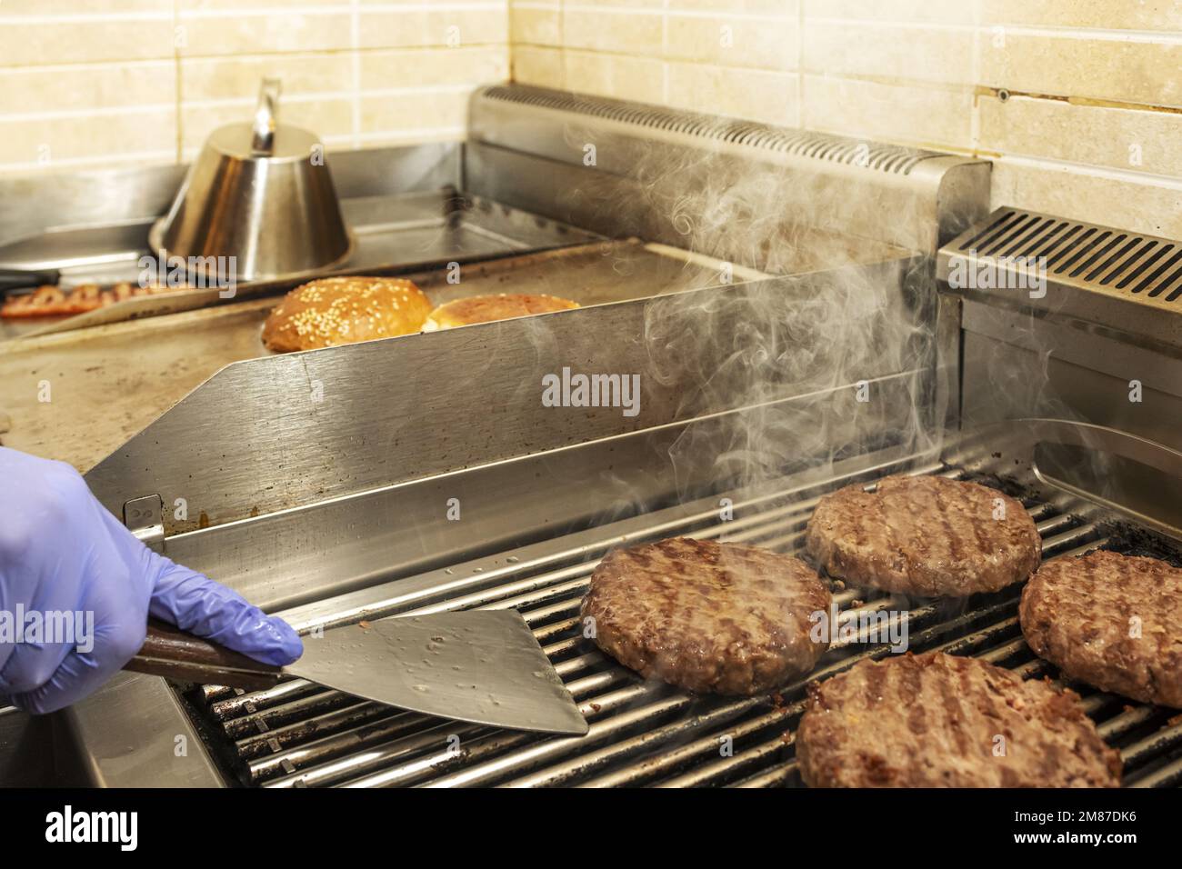A chef with a gloved hand preparing beef burgers on a grill next to ...