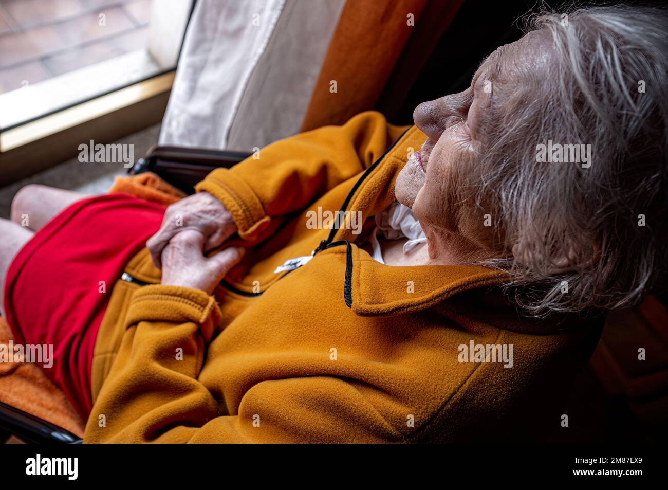 elderly woman in a wheelchair looking out the window with a sad and ...