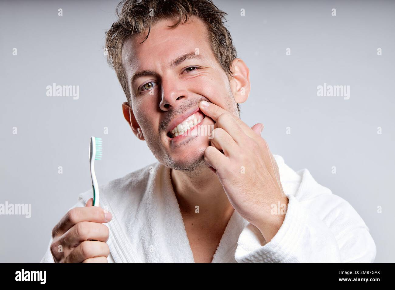 young smiling guy brushing tooth, wearing bathrobe, isolated on gray ...