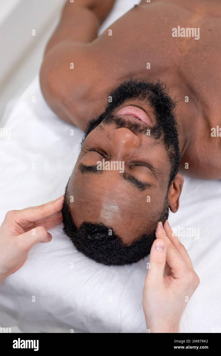 Spa therapist making relaxing massage for handsome middle aged black man. Peaceful bearded man ...