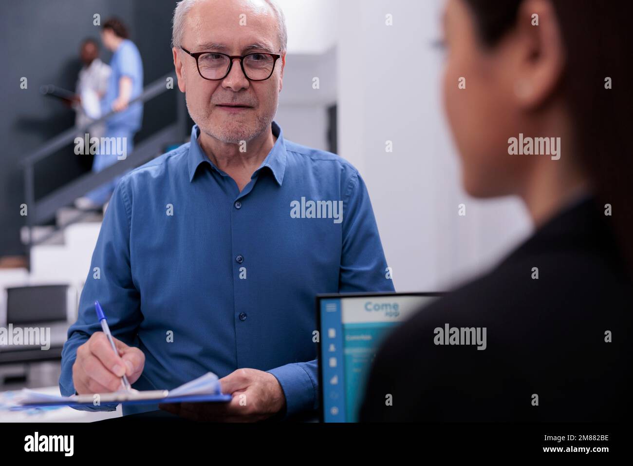Elderly patient standing at hospital counter signing medical documents ...