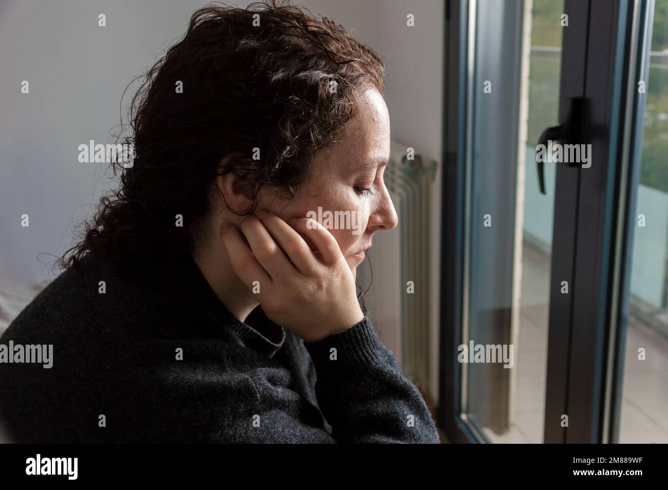 Close-up of sad woman with her head down looking through the window ...