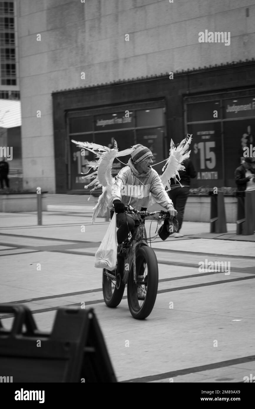 A vertical grayscale of a man riding a bike wearing angel's wings in ...