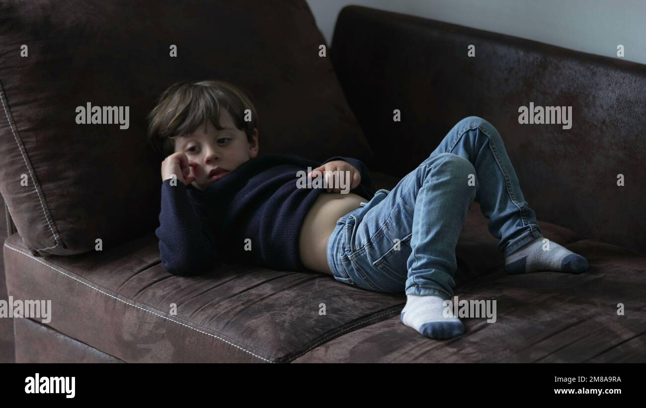 Bored child lying on sofa at home watching TV screen off camera. One ...