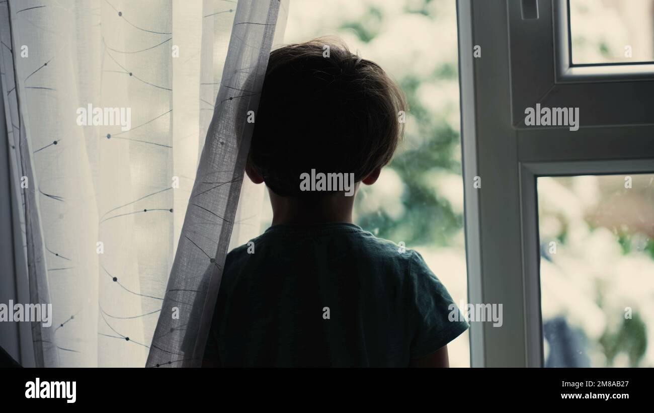 Melancholic little boy standing by window looking through window ...