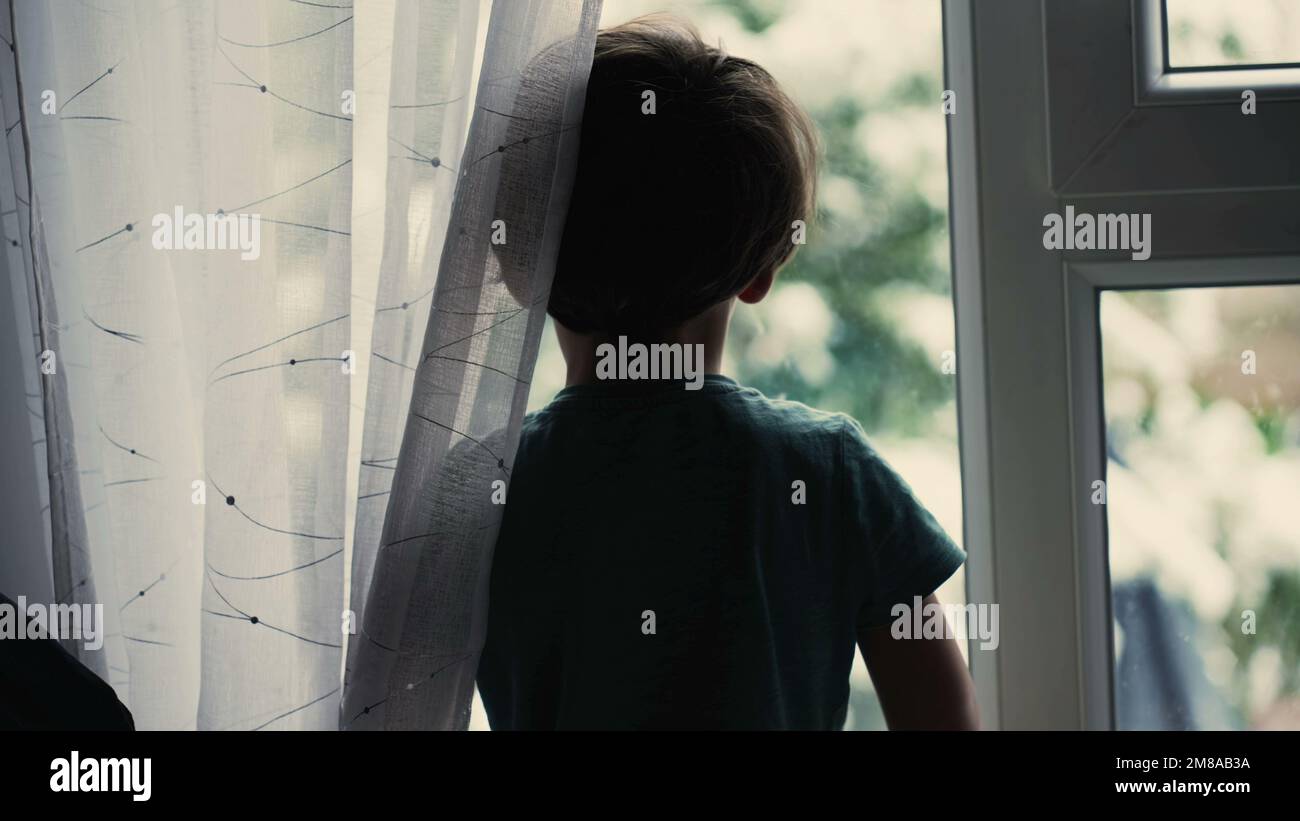 Melancholic little boy standing by window looking through window ...