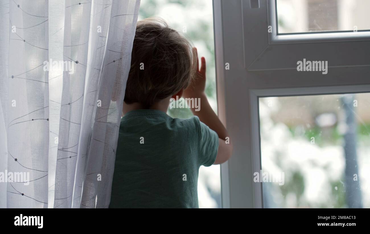 Child standing by window during snowy day. Little boy stands by glass ...
