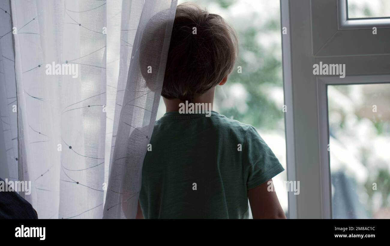 Child standing by window during snowy day. Little boy stands by glass ...