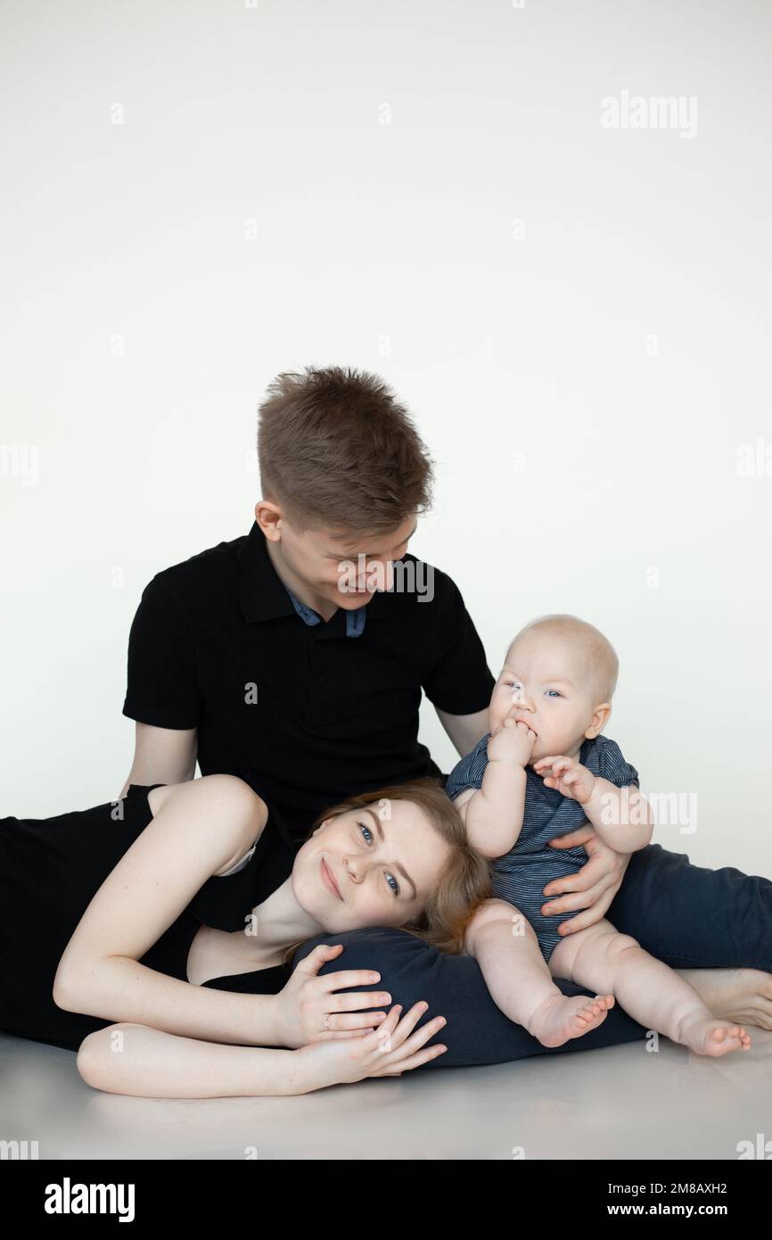 Young family from three in black looks on white background. Dad sit on floor and hold infant ...
