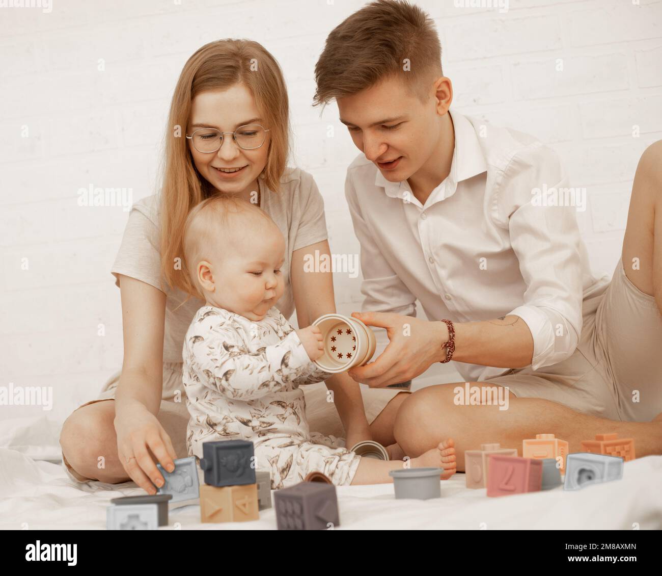 Happy family of three people sit on bed and play toys with little baby ...