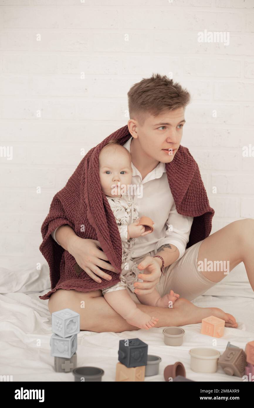 Young father sit under blanket on bed and hug little baby holding in ...