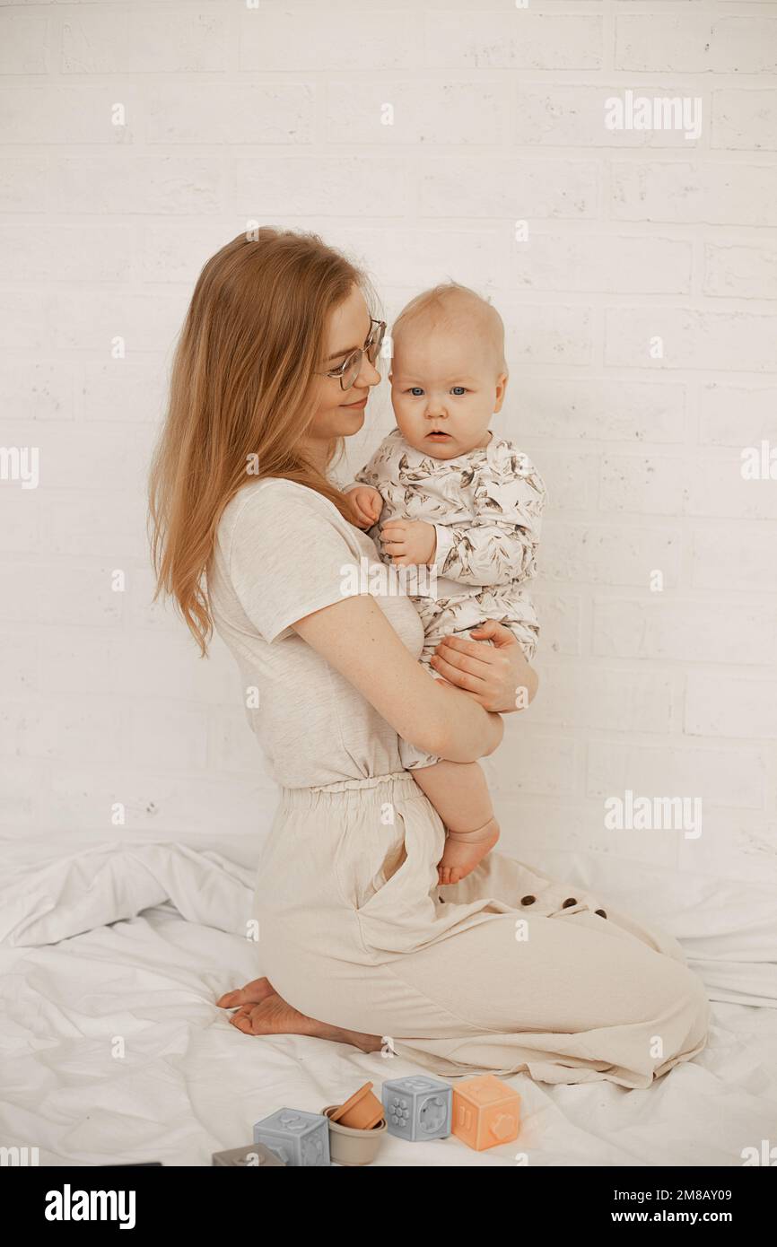 Joyful young mother sit on lap on bed and hold little baby on white background. Home family ...