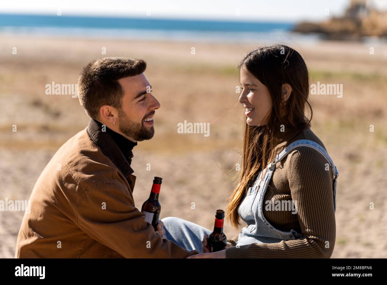 Side view of a cheerful couple with beer bottles smiling and looking at ...