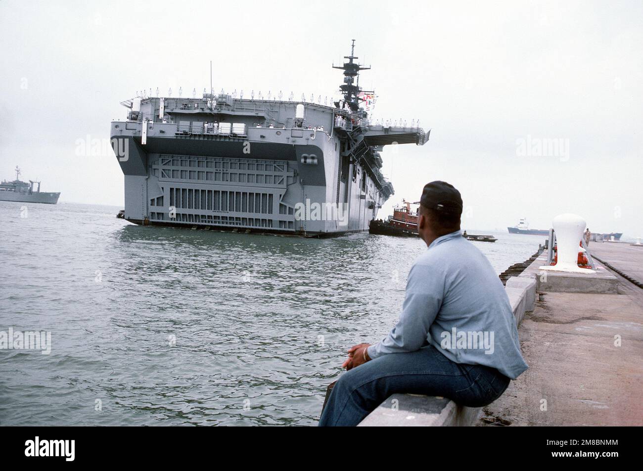 A sailor sitting on a dock faces the stern of the amphibious assault ...
