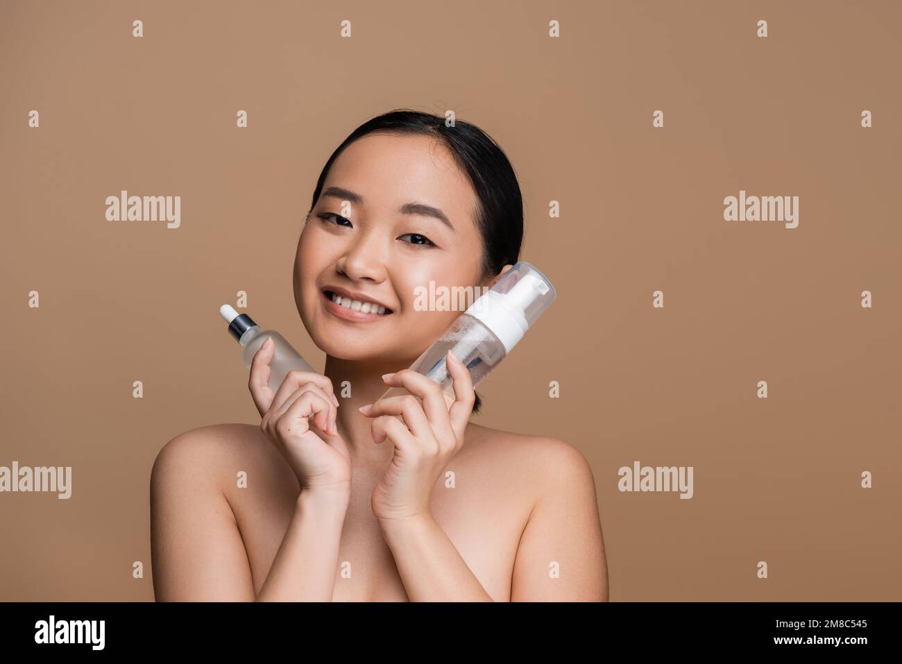 Cheerful asian model holding serum and face foam isolated on brown ...