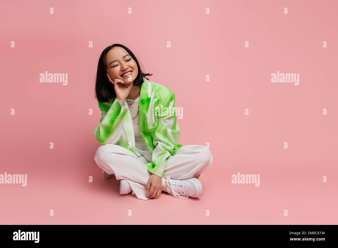 pleased asian woman in fashionable outfit sitting with crossed legs on ...