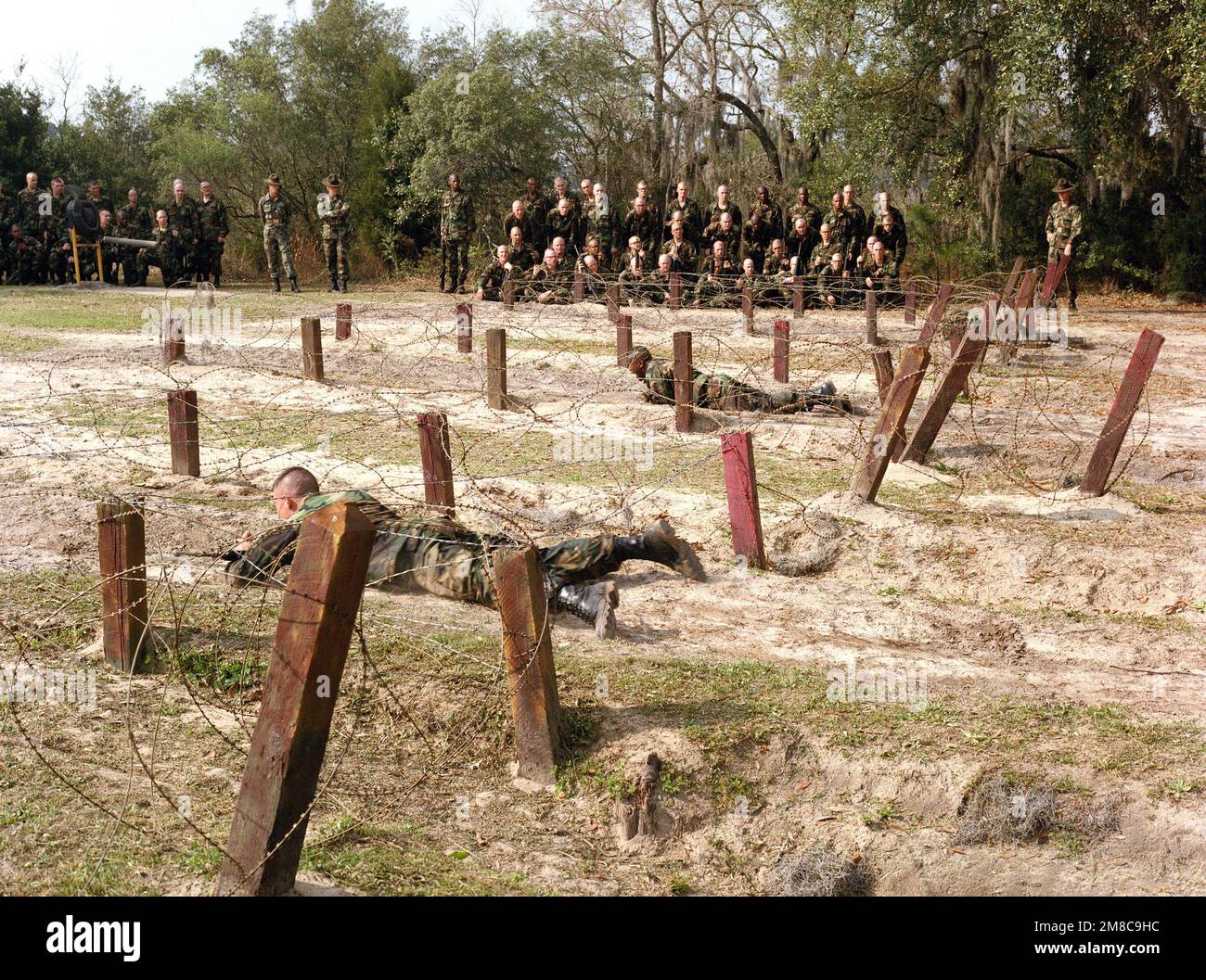 Three drill instructors low crawl under strands of barbed wire to ...