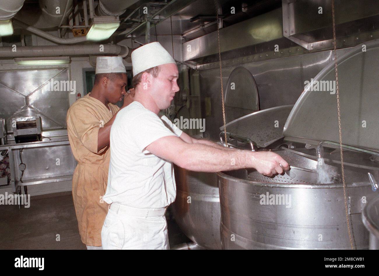 A mess management specialist rinses a colander in the galley of one of ...