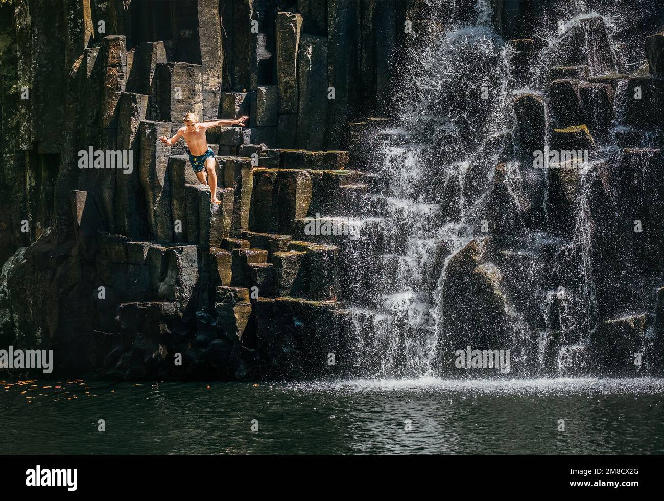 Teenage boy jumping in waterfall lake. Falling water streams flow on ...