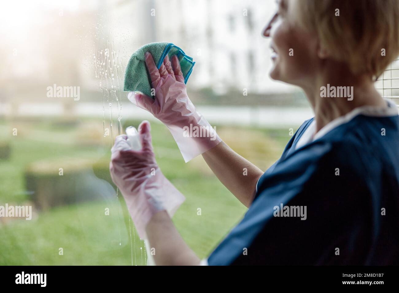 Senior chambermaid in gloves and uniform cleaning window with rag and ...