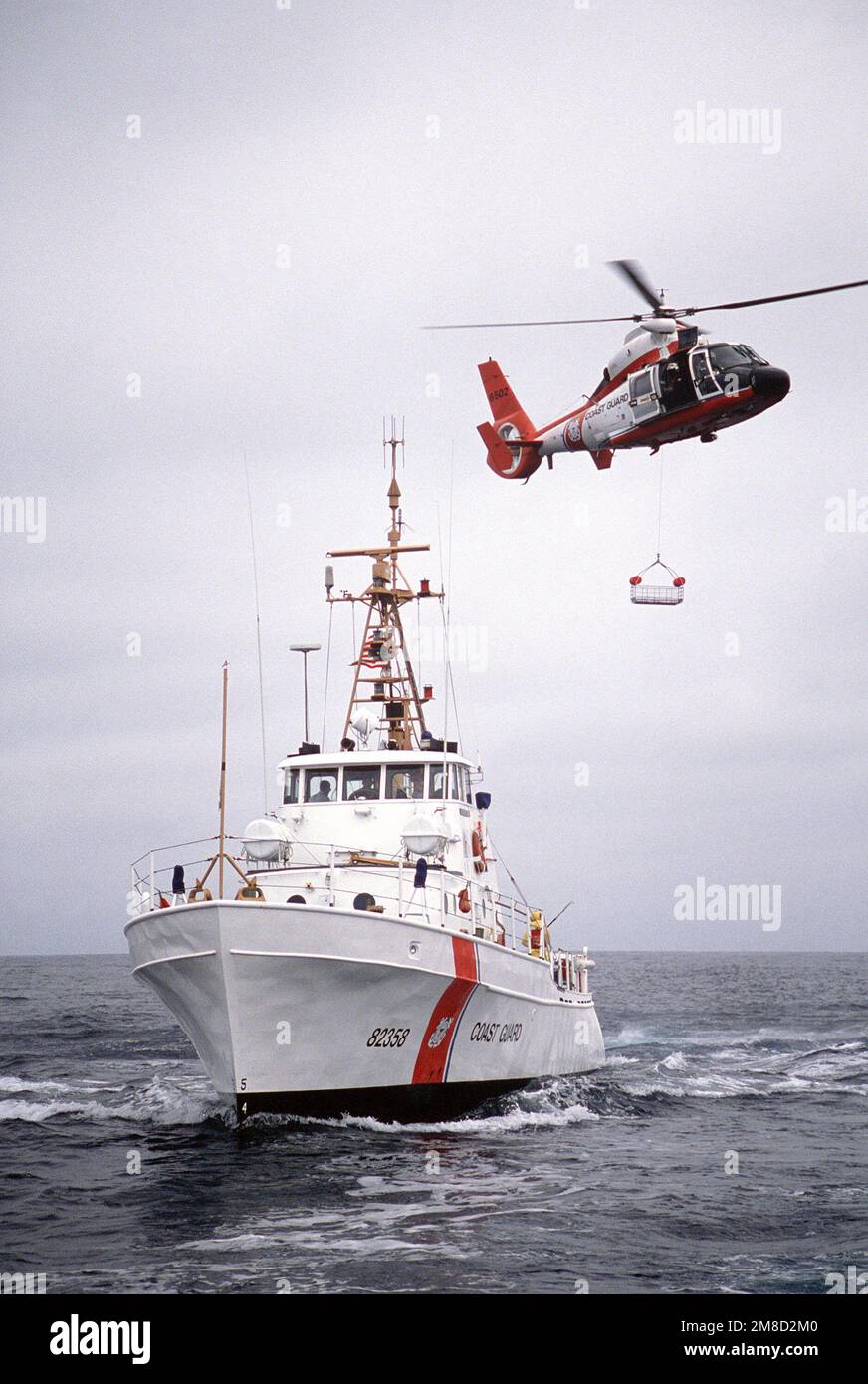 A wire basket is raised toward a US Coast Guard HH-65A Dophin ...