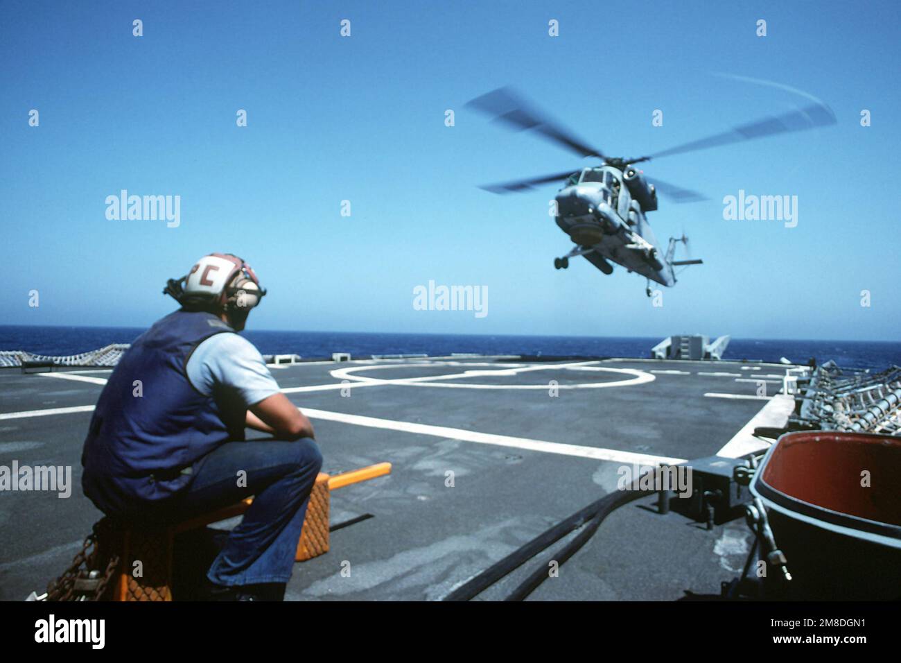 A sailor stands by with wheel chocks and tie-down chains as an SH-2F ...
