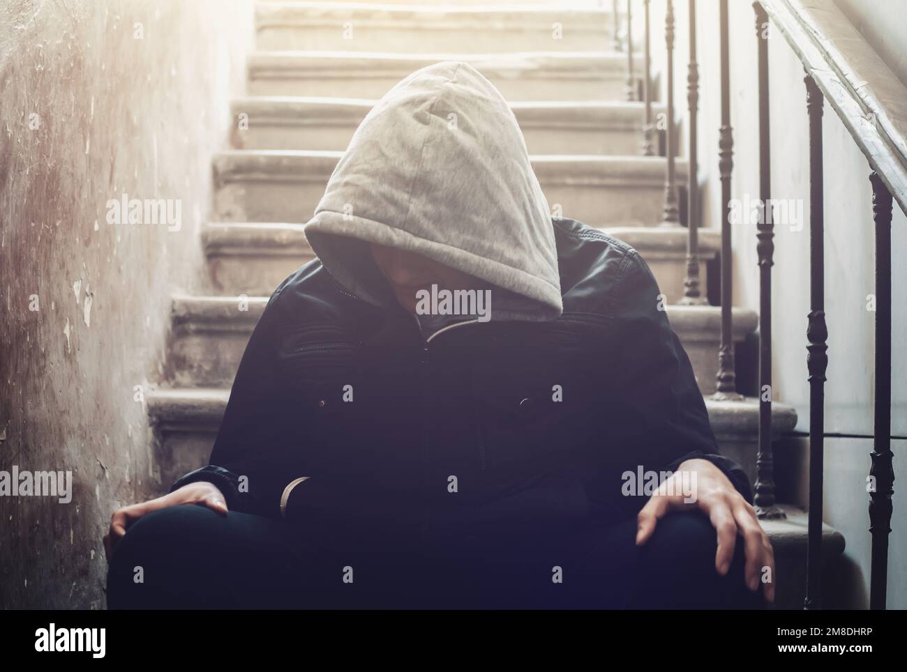 Lonely man in hood with his head down sits on stairs in dark. Stress ...