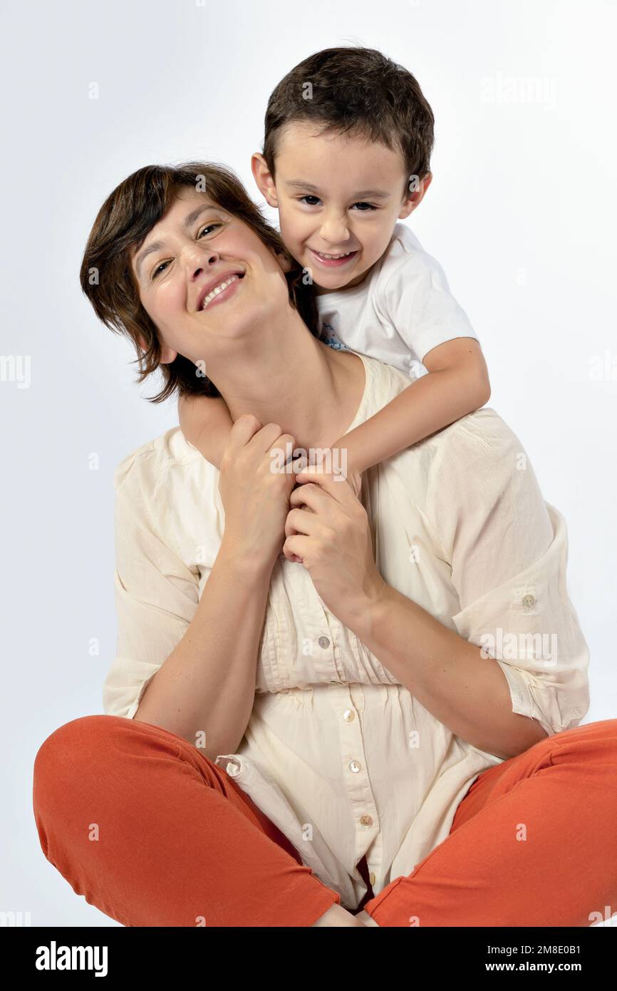 Little boy hugs his mother from behind, both look and smile at camera ...