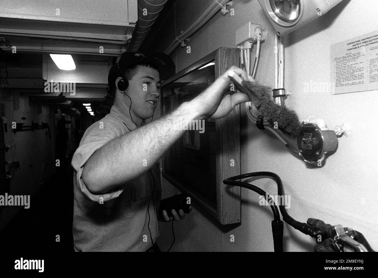 A sailor listens to a tape as he dusts the bulkhead fixtures in a ...