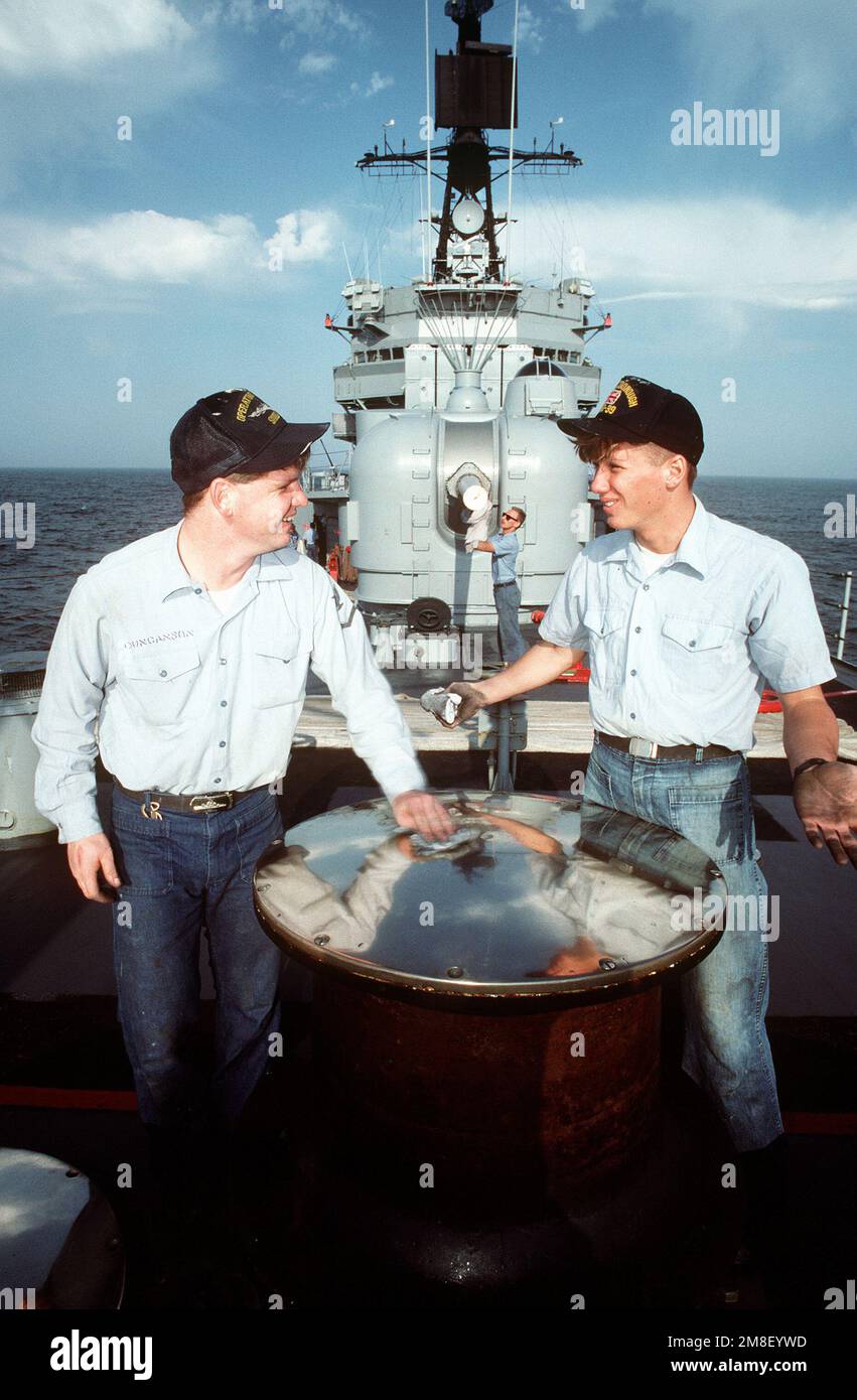 Two sailors talk while polishing the top of a windlass aboard the ...