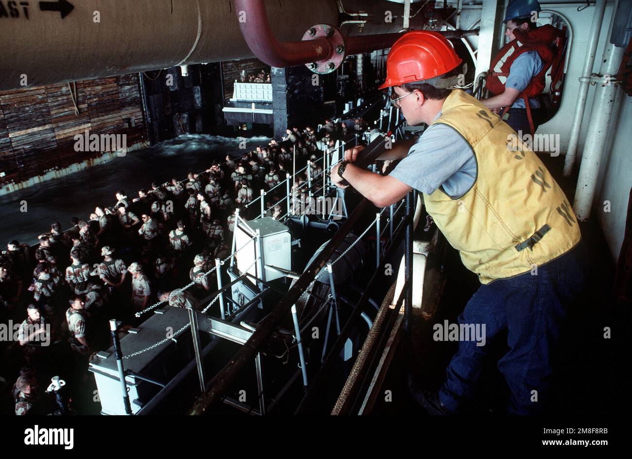 Two sailors watch from a catwalk as a utility landing craft loaded with ...