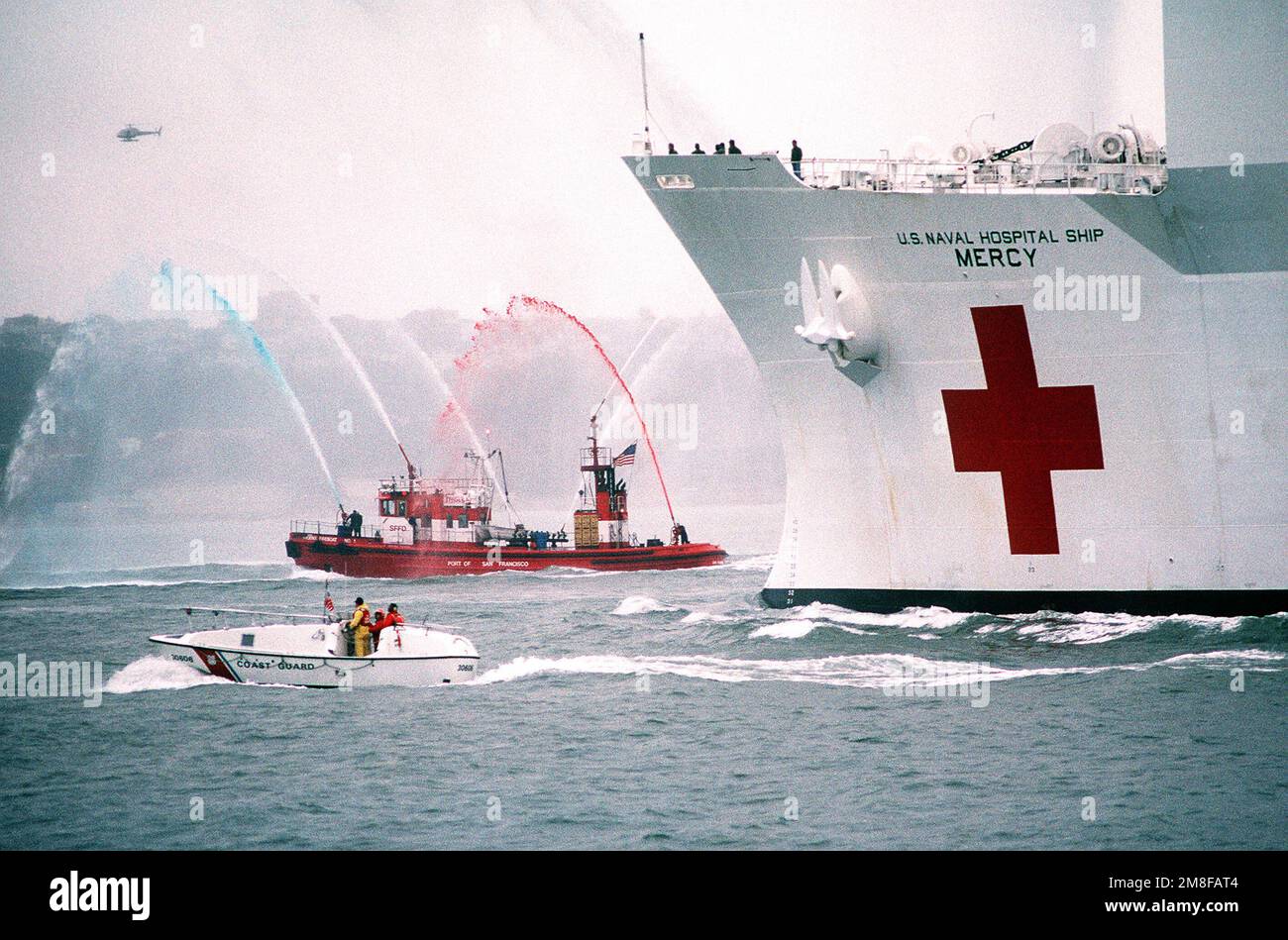 A fire boat sprays water to welcome ship USNS MERCY (T-AH-19) while a U ...