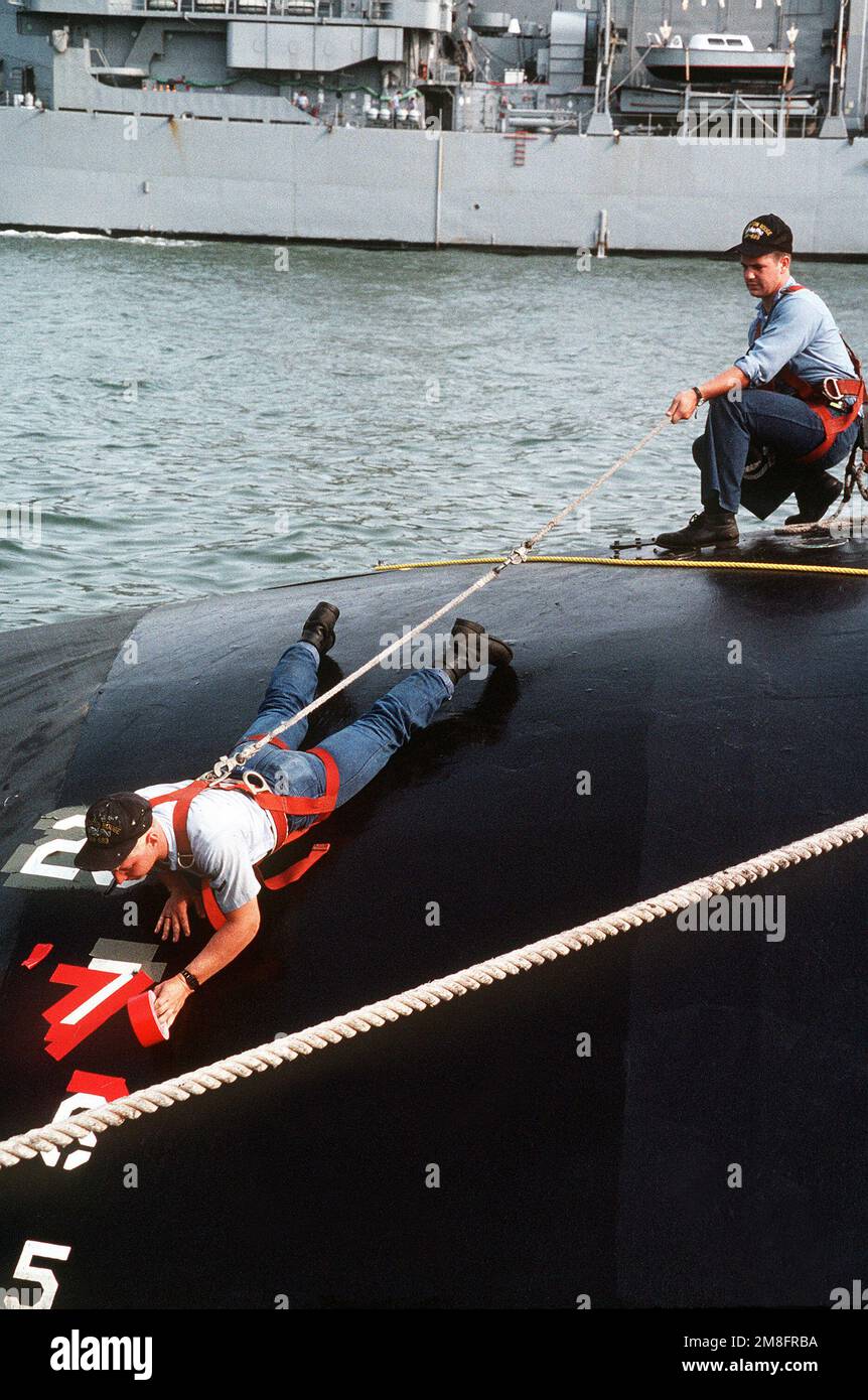 A sailor holds onto the harness as a fellow crew member paints draft ...
