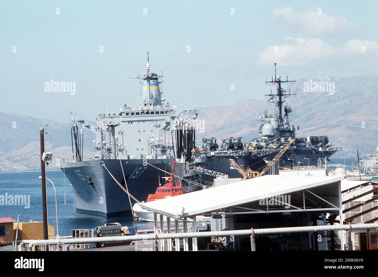 The oiler USNS PECOS (T-AO 197) is tied to a pier, with the amphibious assault ship USS OKINAWA ...