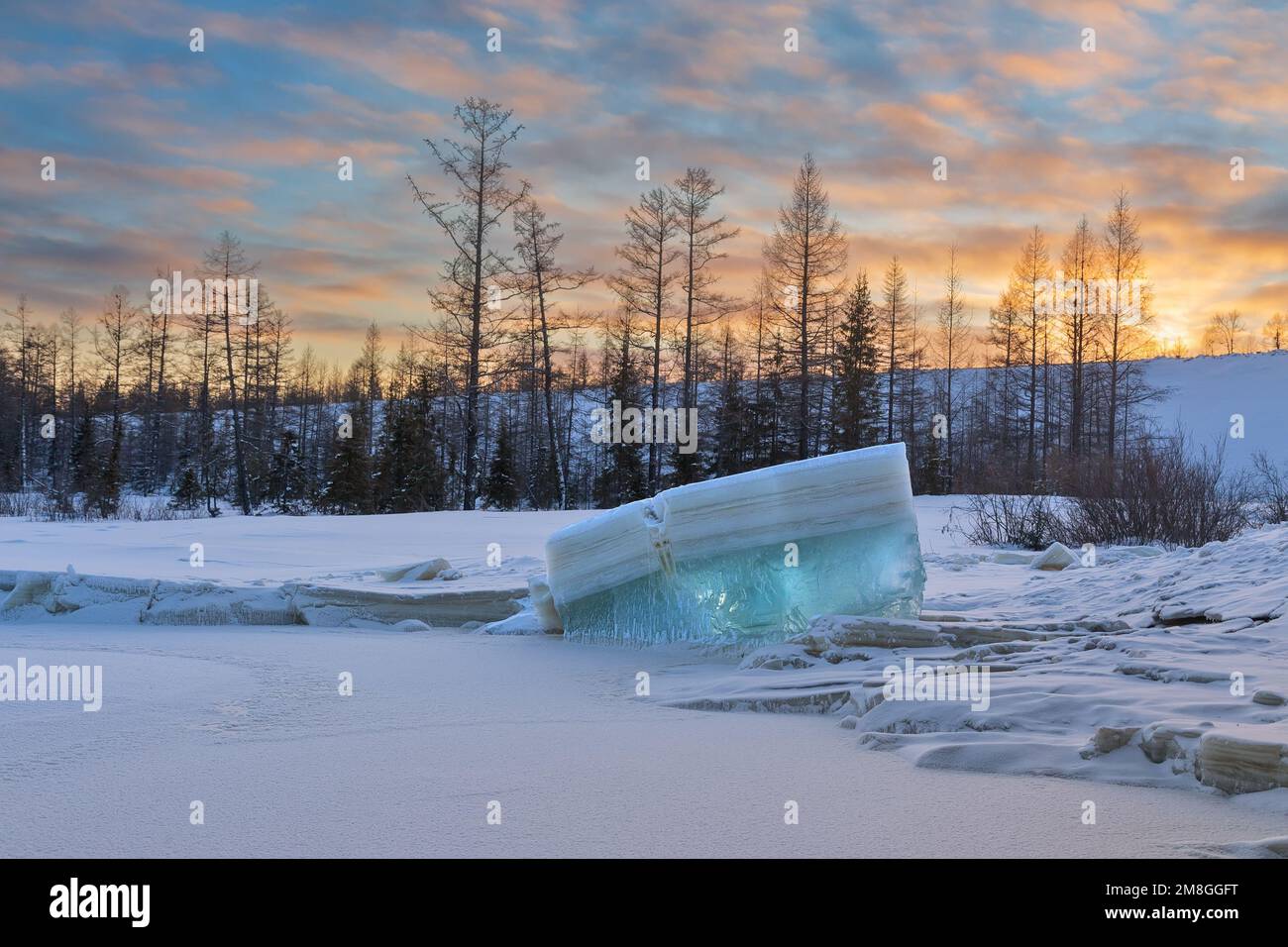 A block of layered turquoise ice on the river bank in the sunset light ...