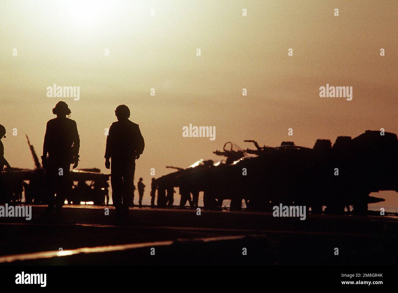 Silhouetted by the setting sun, flight deck crewmen walk toward a row ...